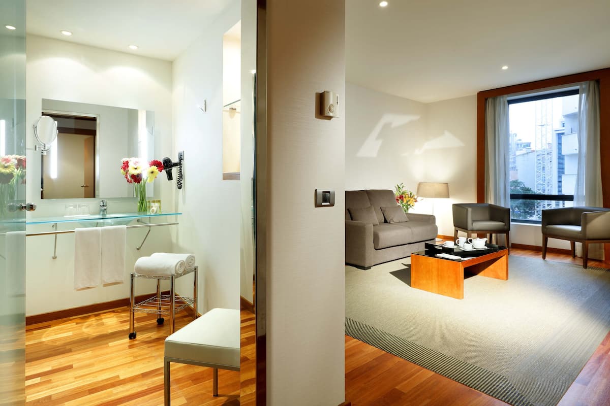 Quarto de hotel moderno com chuveiro de vidro, sofá, mesa de centro e vista da cidade por grandes janelas.