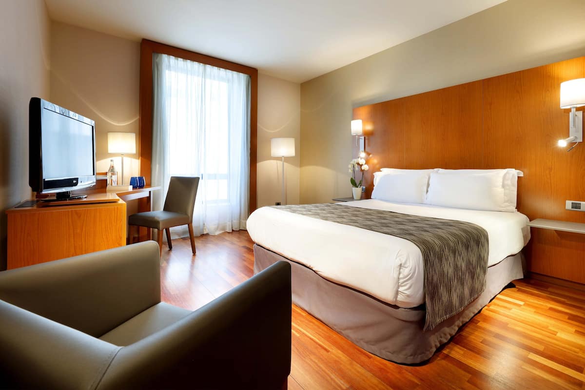 Quarto de hotel com cama, escrivaninha com televisão, uma cadeira e abajur.