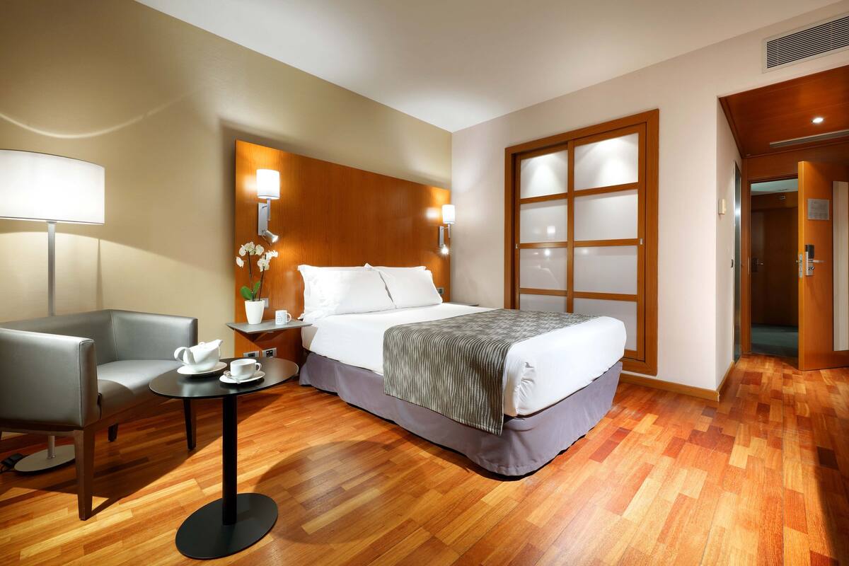 Quarto de hotel com uma cama, uma cadeira, uma mesinha de centro com uma luminária e uma mesa de cabeceira com uma luminária e um vaso de flores.