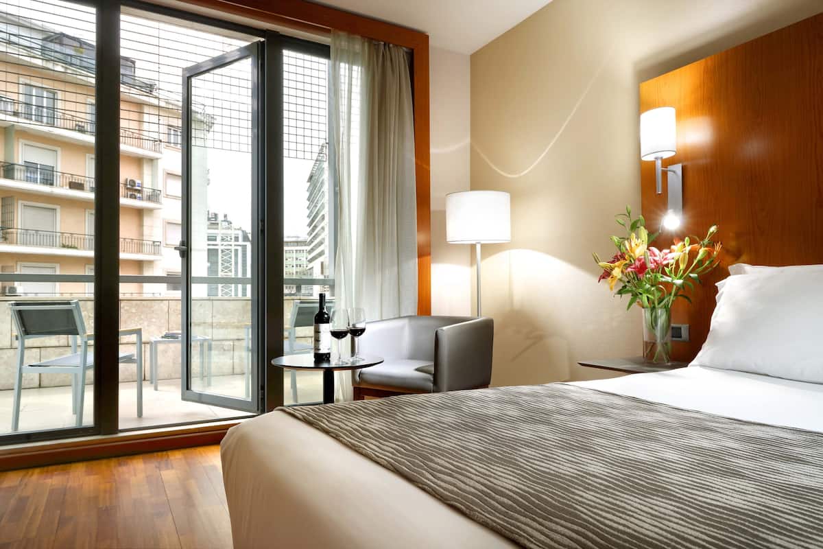 Quarto de hotel com cama, escrivaninha com cadeira, abajur e um vaso com flores.