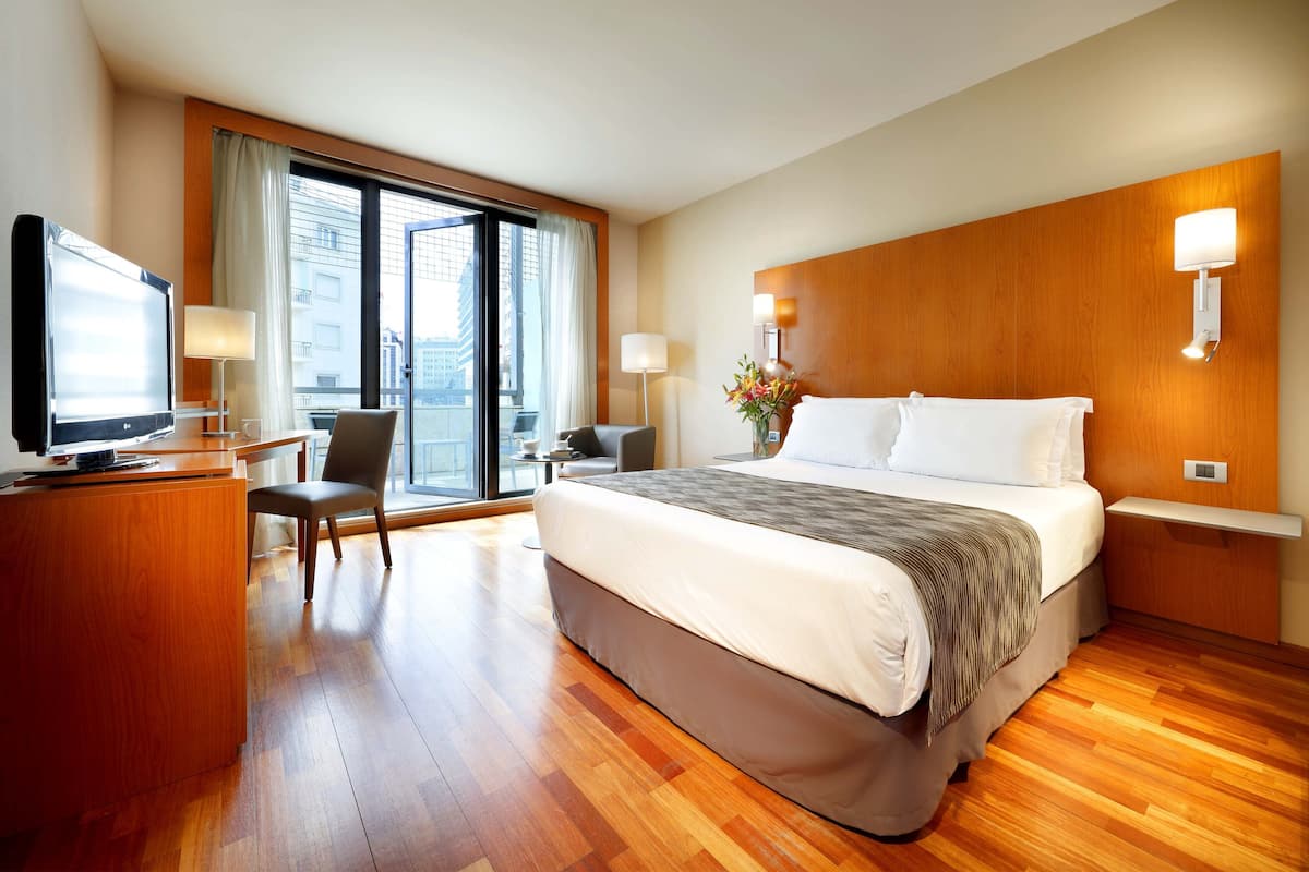 Quarto de hotel com uma cama grande, uma escrivaninha com televisão, uma cadeira, uma luminária de mesa, uma mesinha de cabeceira com um vaso de flores e vista para a cidade pela janela.