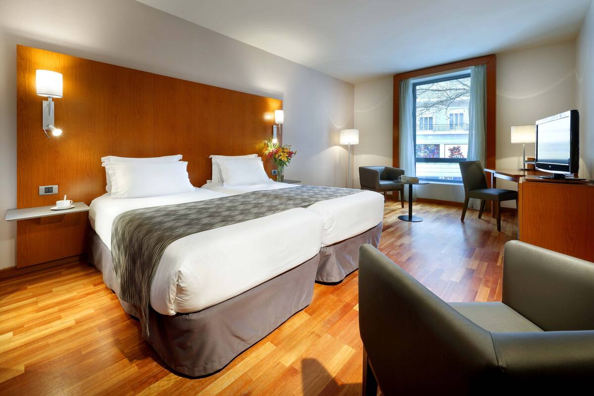 Quarto de hotel com uma cama grande, uma escrivaninha com cadeira, televisão e janela com vista para edifícios.