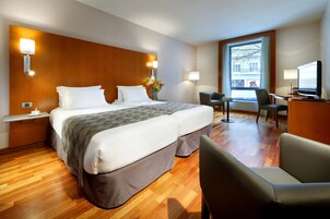 Quarto de hotel com uma cama grande, uma escrivaninha com cadeira, televisão e janela com vista para edifícios.