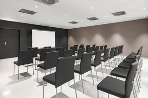 Uma sala de conferência com fileiras de cadeiras pretas, uma tela de projeção branca e uma parede bege.