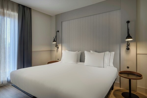 Uma cama bem arrumada com lençóis brancos, duas mesas de cabeceira com abajures e uma janela grande com cortinas.