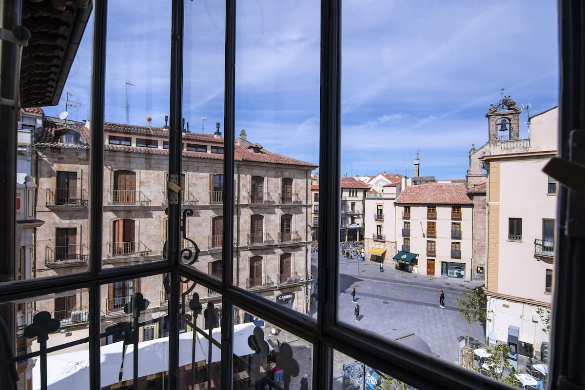 Visão de uma praça histórica, com edifícios tradicionais, varandas e um campanário.