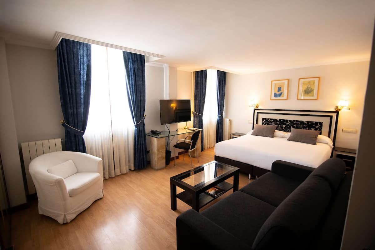 Quarto de hotel com cama, sofá, escrivaninha com televisão, cadeira e cortinas.