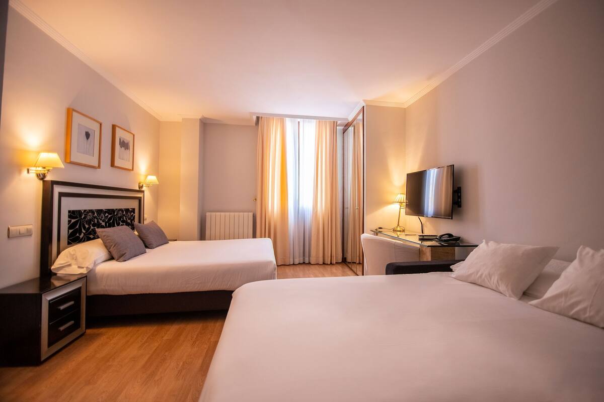 Quarto de hotel com duas camas, televisão, abajures, escrivaninha e janela com cortinas.