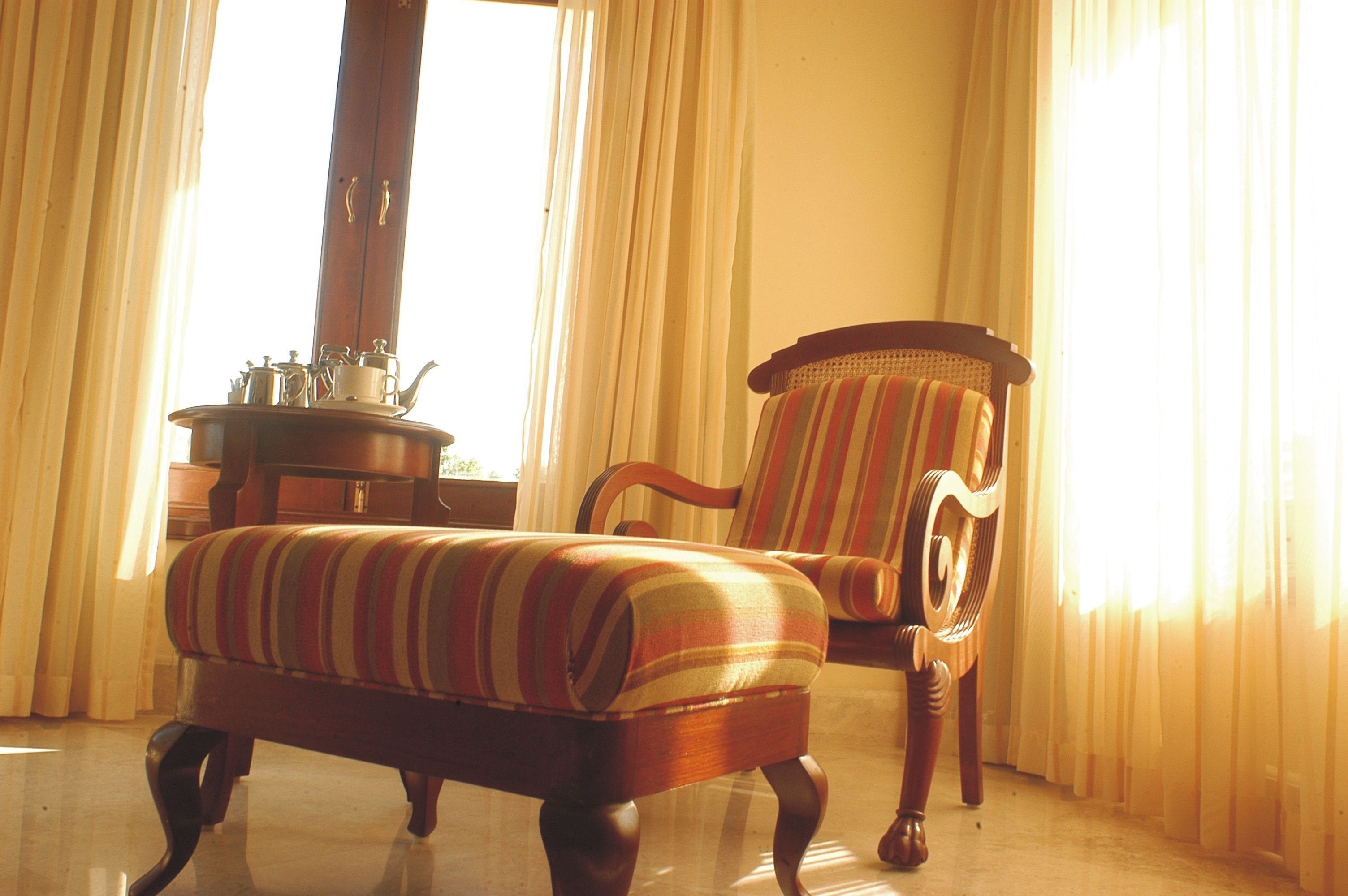 Quarto com uma cadeira de madeira, um banquinho listrado e uma mesa com um bule.
