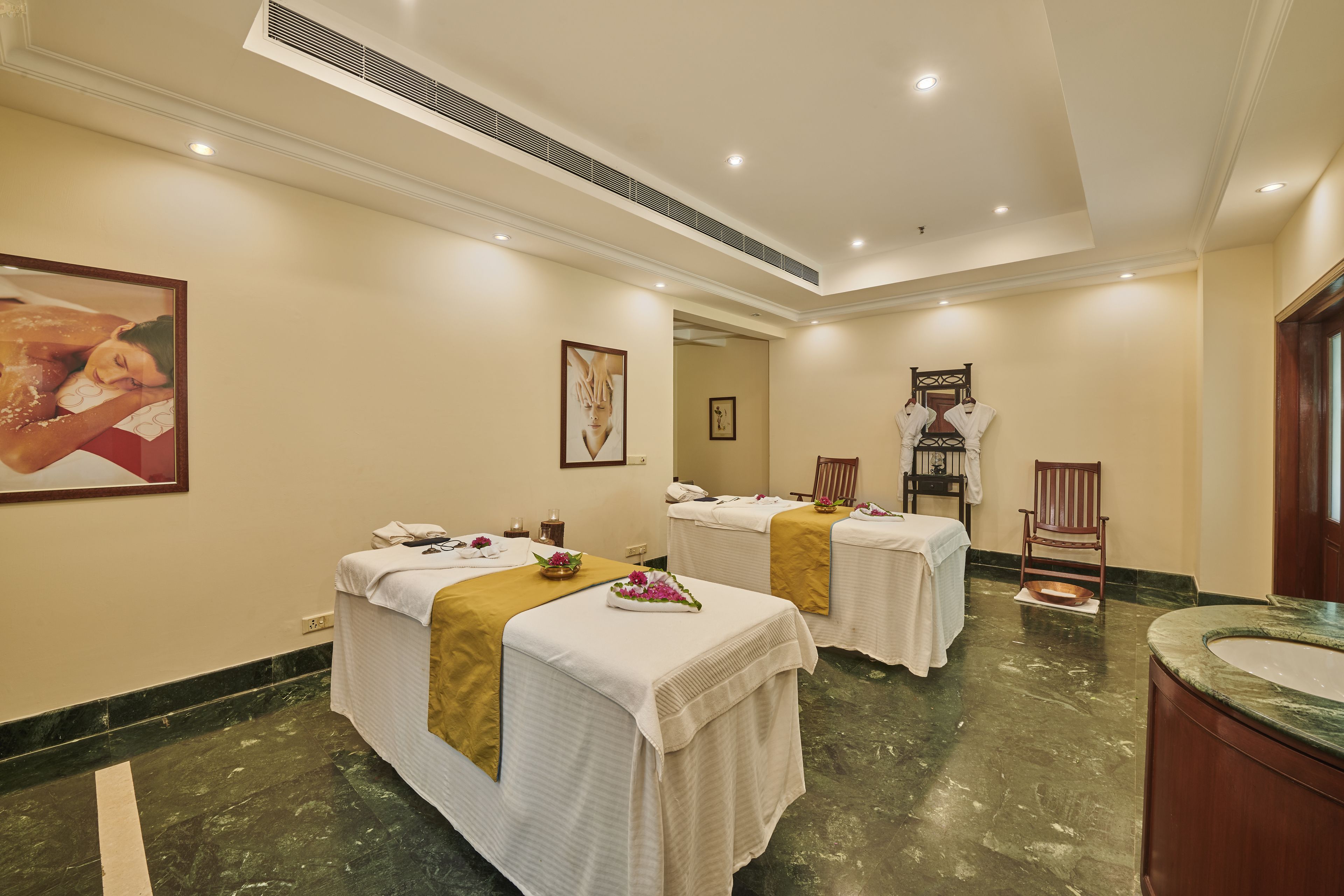 Um espaço de spa com duas mesas de massagem, uma cadeira de madeira, piso de mármore e um espelho grande.