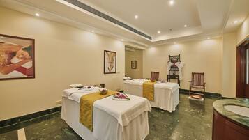 Um espaço de spa com duas mesas de massagem, uma cadeira de madeira, piso de mármore e um espelho grande.