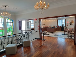 Um hall de hotel com piso de madeira, uma escada com guarda-corpo em ferro forjado, um lustre e vista para um jardim através de uma janela com vitrais.