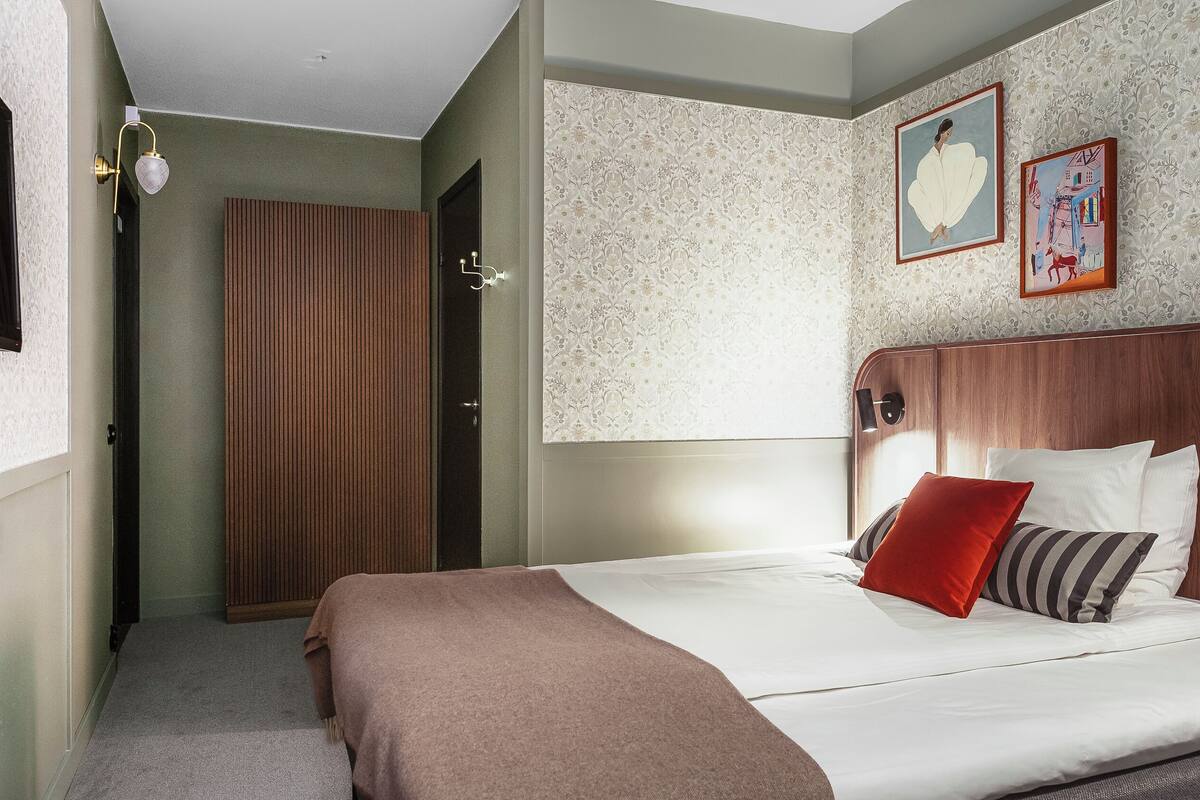 Quarto de hotel com uma cama, cabeceira de madeira, porta e parede com papel de parede estampado e obras de arte.