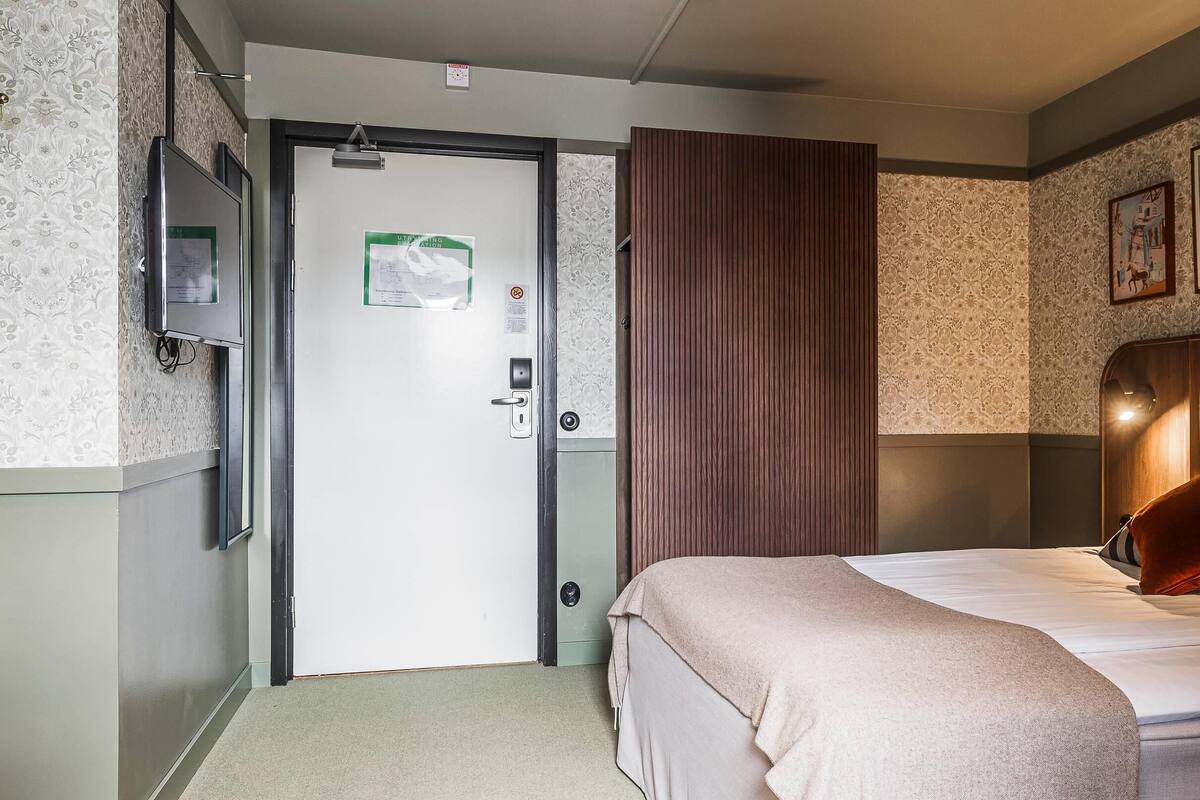 Quarto de hotel com cama, porta com câmera de segurança, televisão na parede e quadro decorativo.