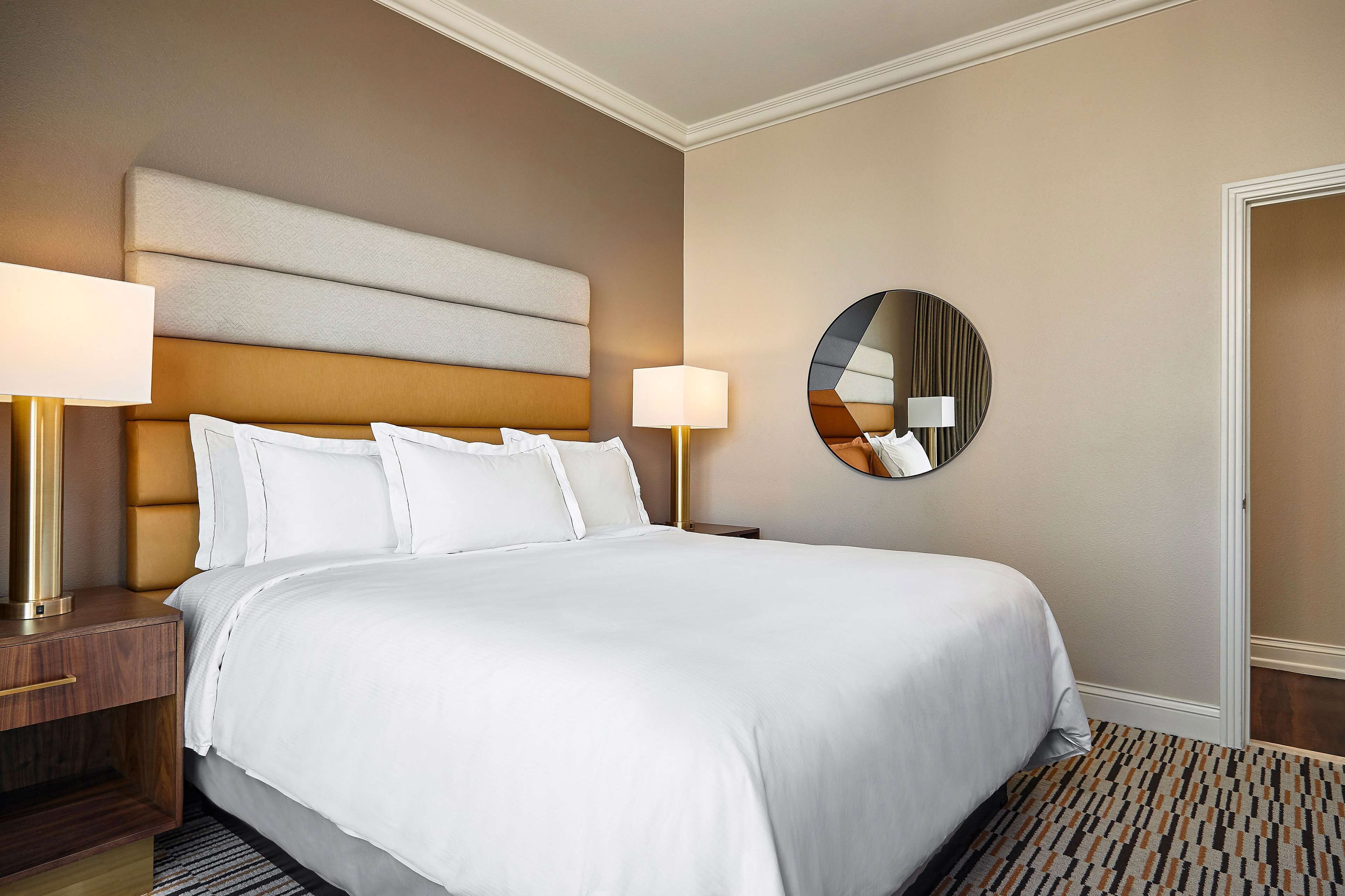 Quarto de hotel com uma cama grande, duas mesas de cabeceira com abajures, um espelho redondo e um tapete estampado.