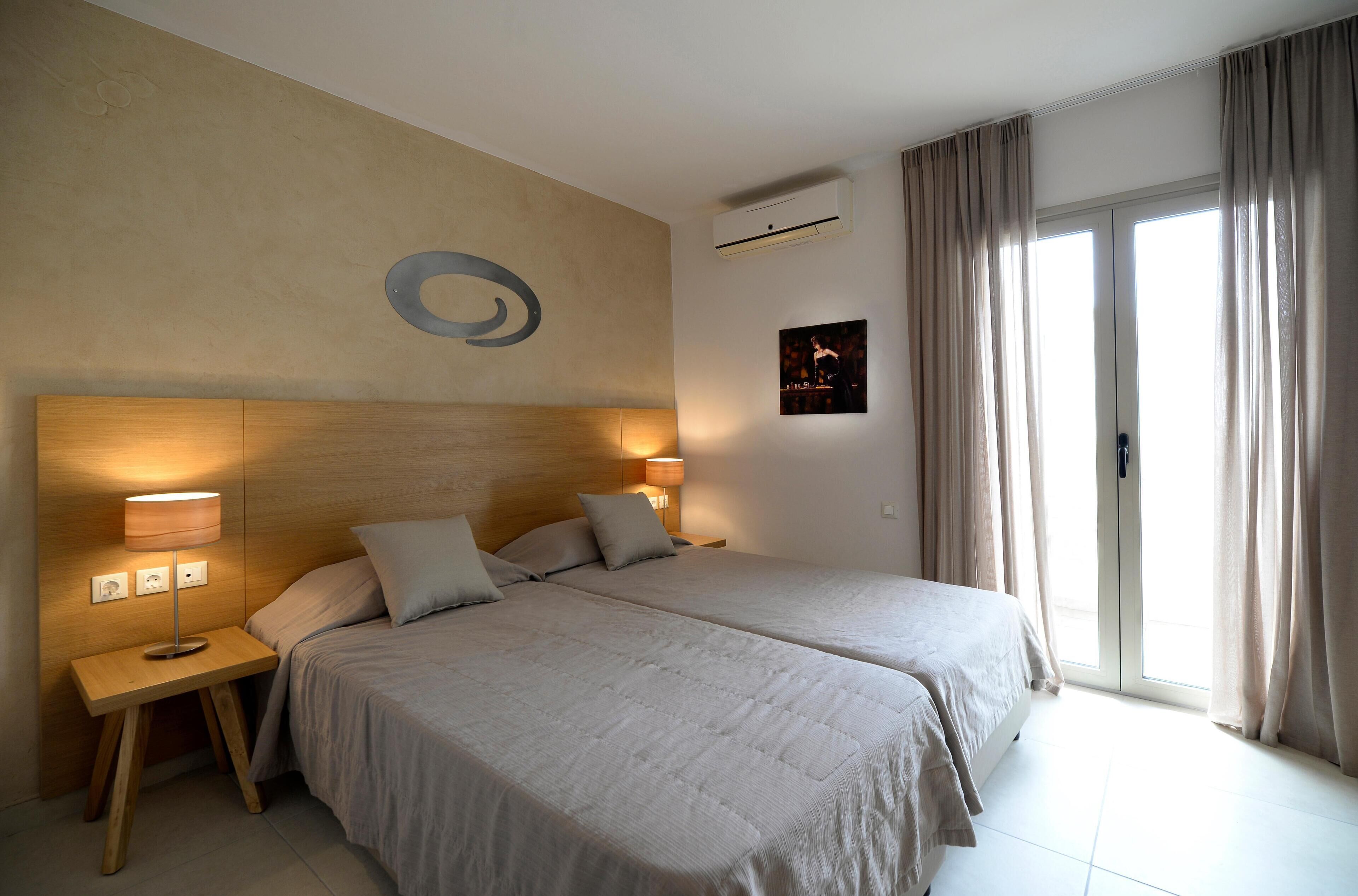 Uma cama de casal com cabeceira de madeira, duas mesas de cabeceira com abajures, um ar condicionado fixado na parede e uma janela grande com cortinas.