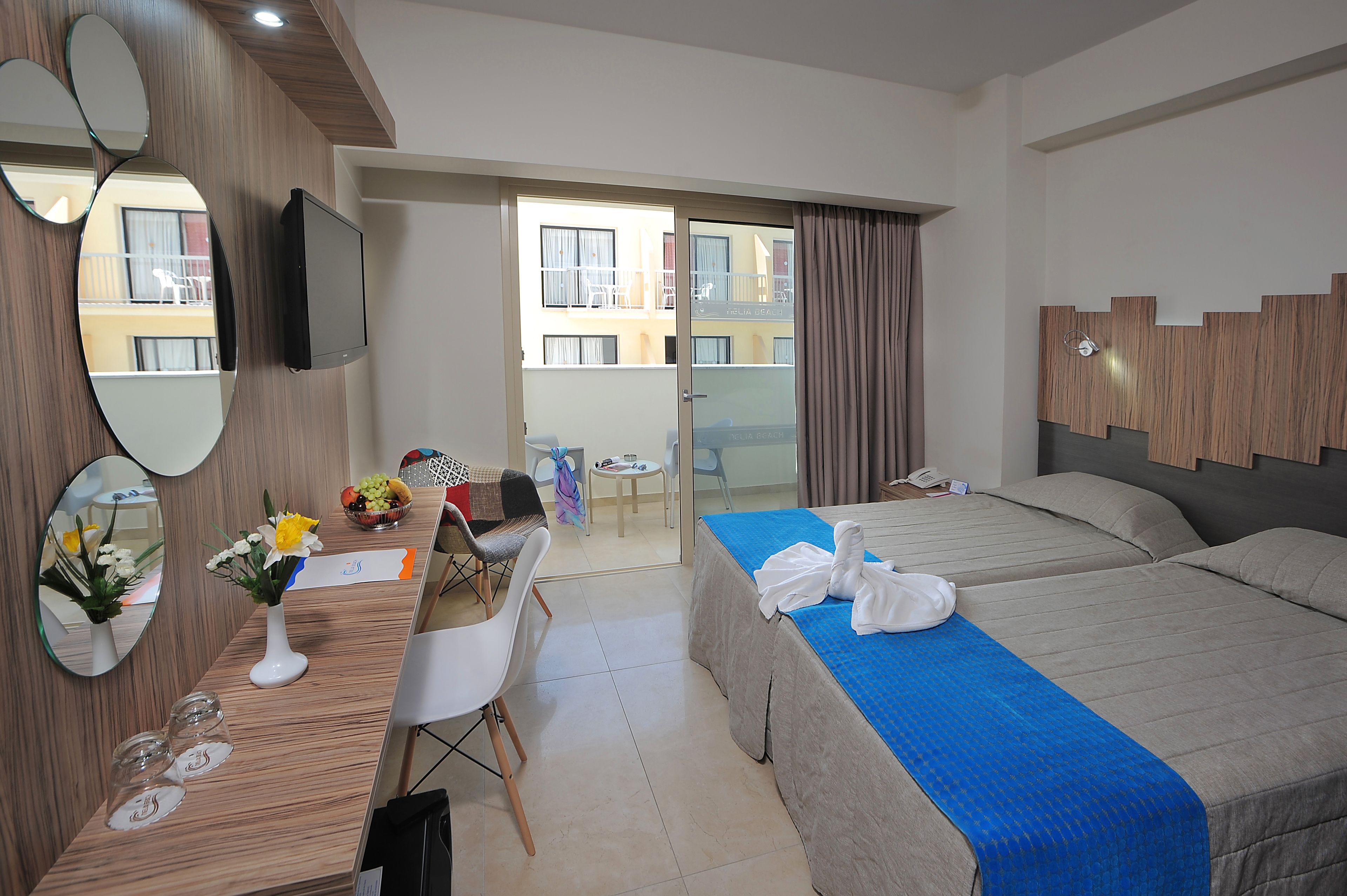 Quarto de hotel com duas camas, uma escrivaninha com cadeira, um espelho e uma varanda com vista para o edifício.