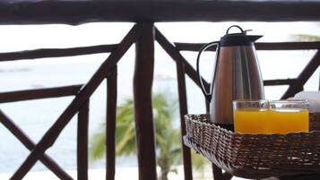 Uma cesta de vime com uma garrafa de água de aço inoxidável e dois copos de suco de laranja numa varanda com vista para o mar.