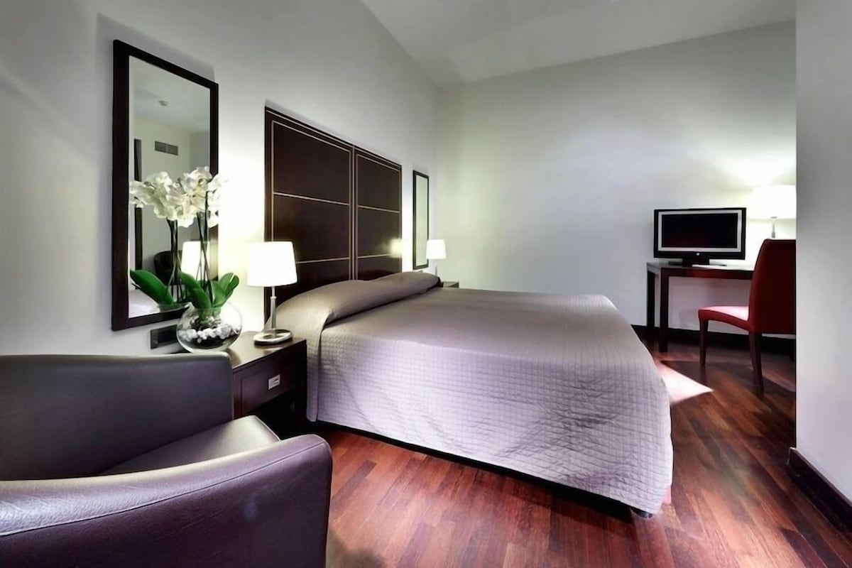 Quarto de hotel com uma cama, escrivaninha, cadeira, televisão e espelho.