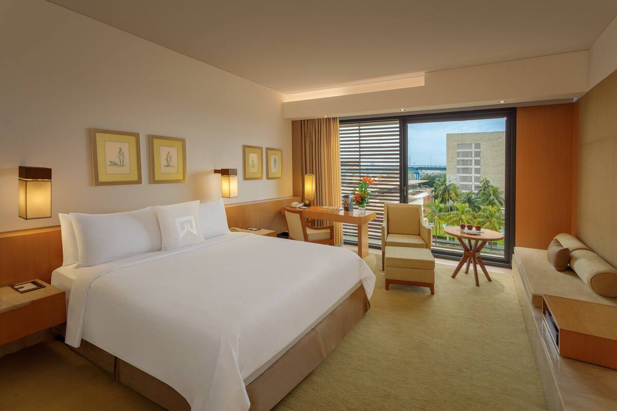 Quarto de hotel com uma cama grande, escrivaninha, cadeira, sofá e varanda com vista para palmeiras.