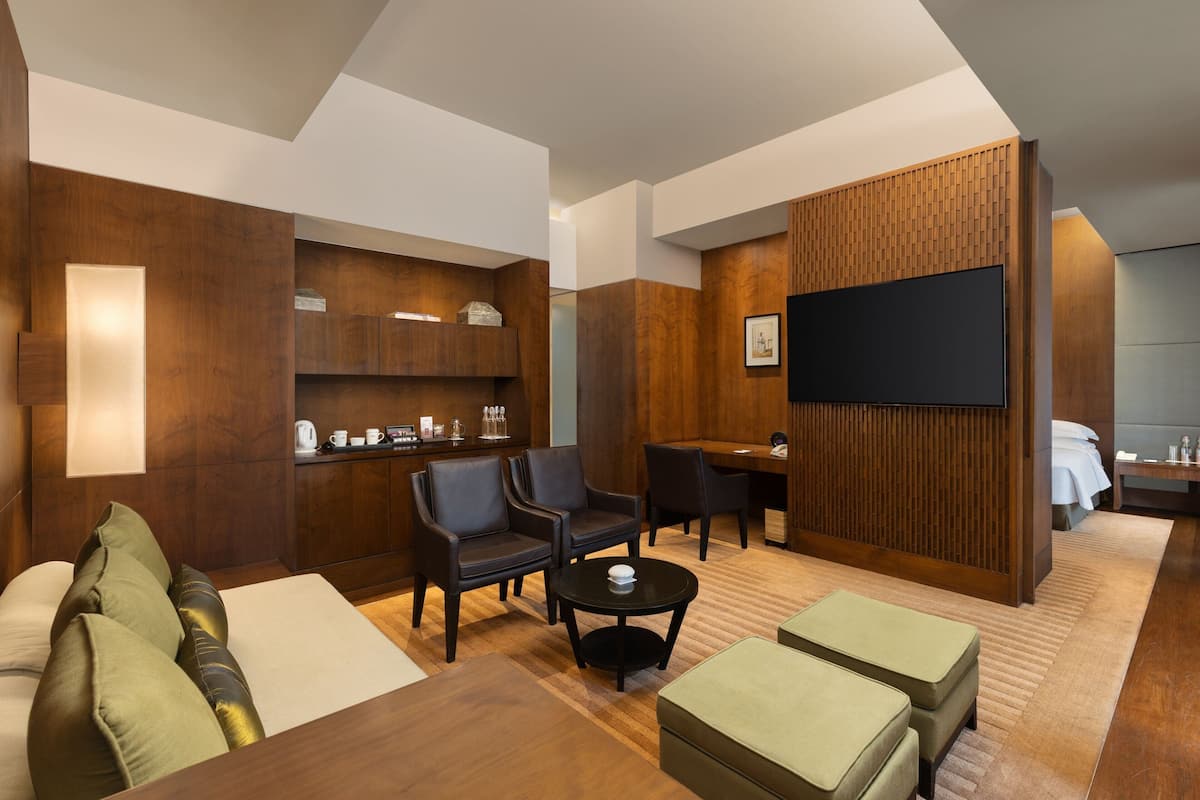 Quarto de hotel moderno com parede de destaque em madeira, televisão de tela plana, sofá, mesa de centro e área de refeições.