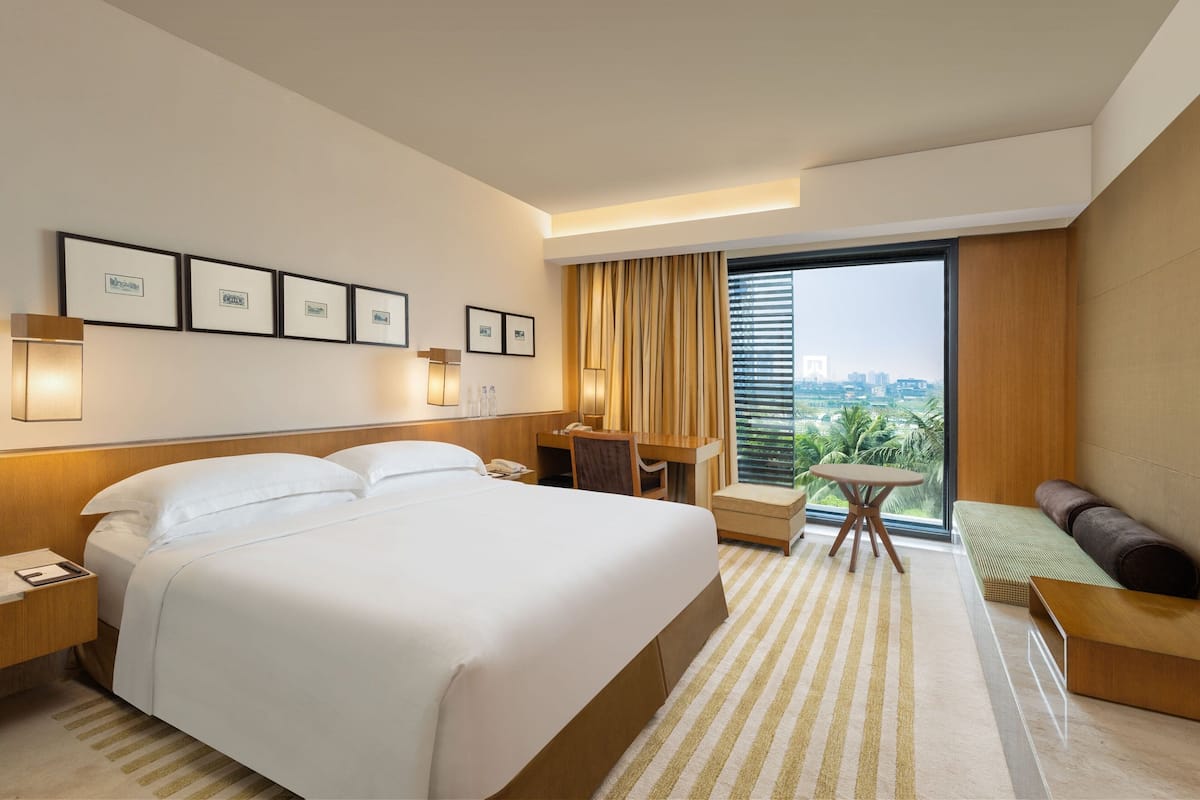Quarto de hotel com uma cama grande, uma escrivaninha, uma cadeira, uma mesa pequena e um sofá. Há quadros nas paredes e uma janela com vista para a cidade.