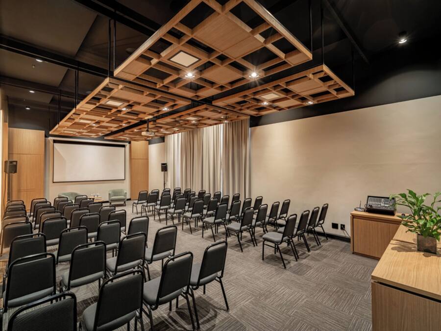 Uma sala de conferências com fileiras de cadeiras, um pódio, uma tela de projetor e um design de teto em madeira.