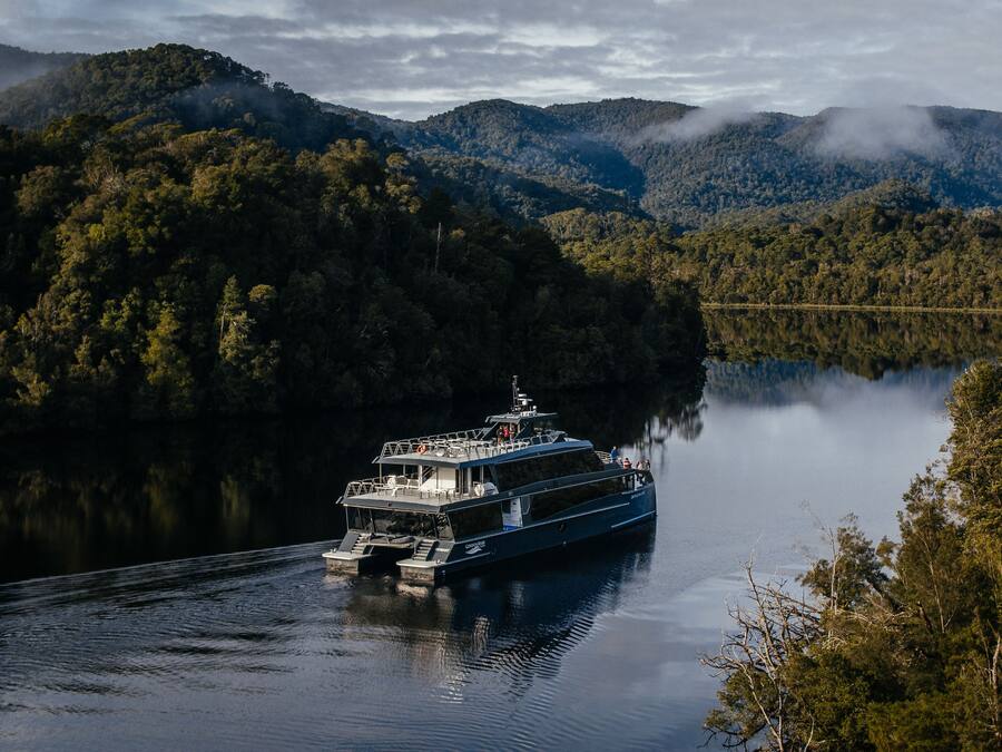Um barco de passeio flutuando em um rio calmo, cercado por floresta densa e montanhas.