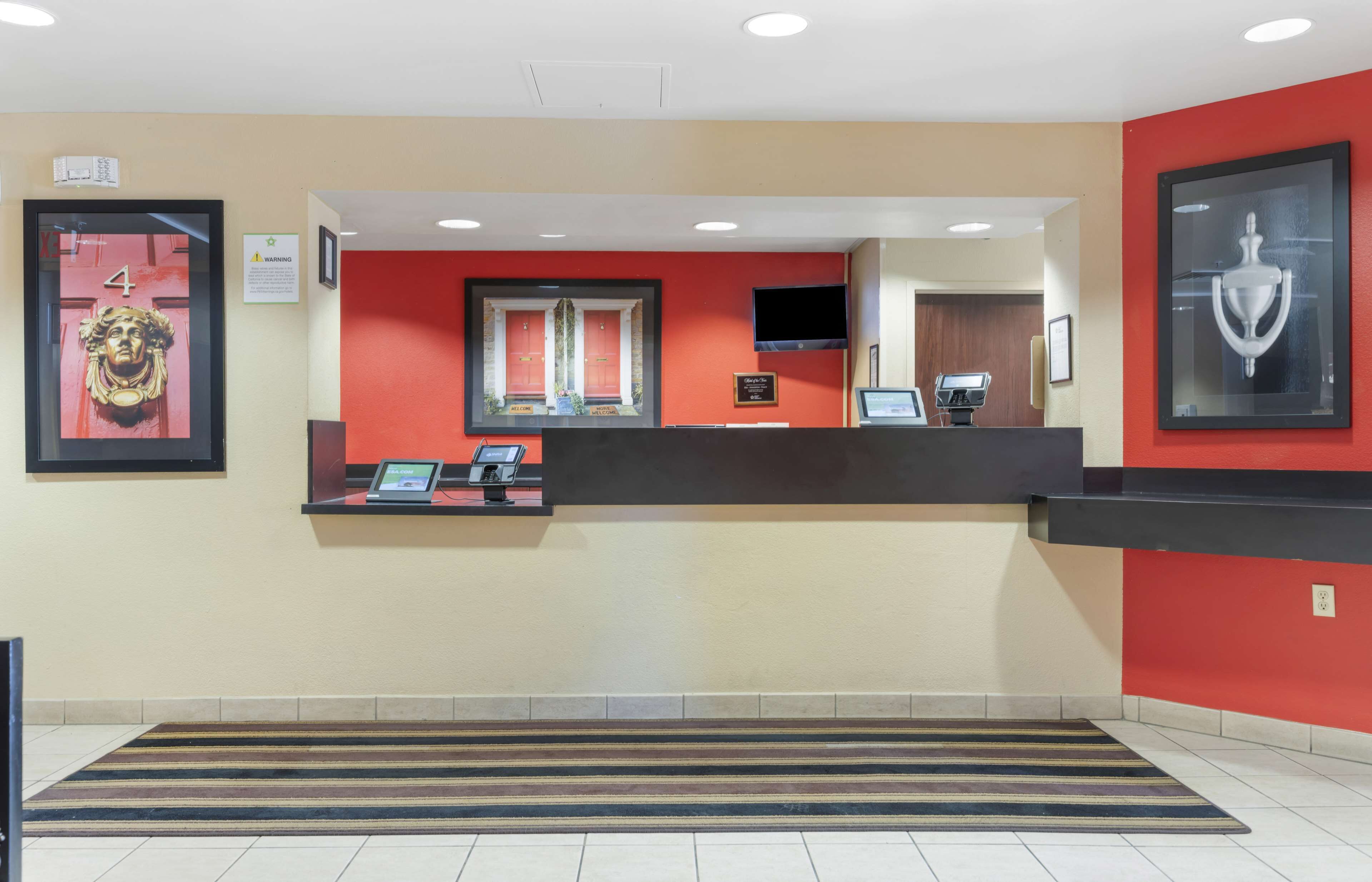 Uma área de recepção de hotel, com balcão de atendimento, um quiosque de check-in digital e quadros nas paredes.
