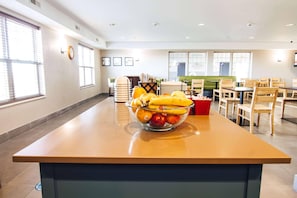 Um saguão de hotel com uma ilha central, com uma cesta de frutas, área de estar com mesas e cadeiras, e janelas amplas que proporcionam luz natural.