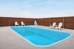 Uma piscina retangular de cor azul clara, cercada por uma cerca de madeira. Quatro cadeiras de plástico branco estão dispostas ao redor da piscina.