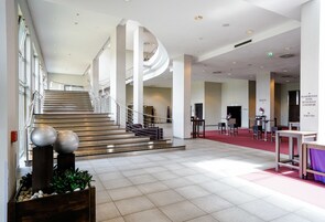 Um hall de hotel espaçoso com uma escadaria curva, área de recepção e piso coberto por tapete vermelho.