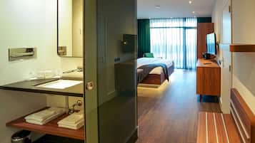 Quarto de hotel moderno com banheiro envidraçado, cama com cabeceira verde, escrivaninha de madeira e uma janela grande com cortinas.