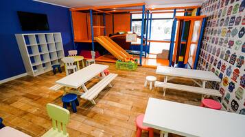 Um espaço de brincadeiras infantil com escorrega, mesas, cadeiras e uma parede decorada com personagens de desenho animado.