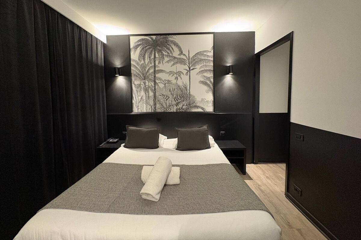 Quarto de hotel moderno com uma cama grande, uma cortina escura, uma obra de arte na parede com palmeiras, duas mesas de cabeceira com abajures e um espelho grande.
