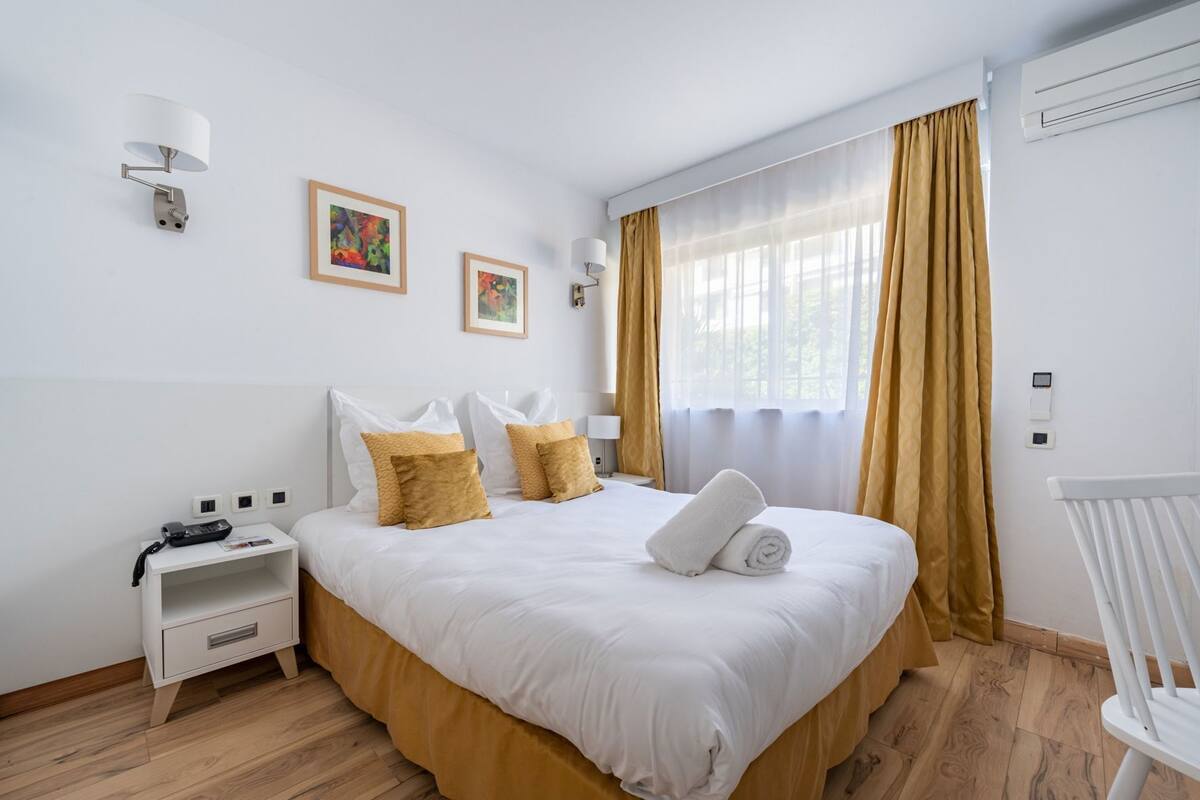 Quarto de hotel com cama, mesas de cabeceira, uma cadeira, janela com cortinas e luminárias fixadas na parede.