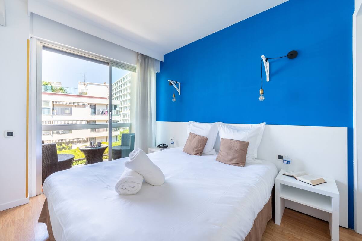 Quarto de hotel com uma cama grande, uma mesinha de cabeceira, uma janela com vista para edifícios e uma parede azul de destaque.