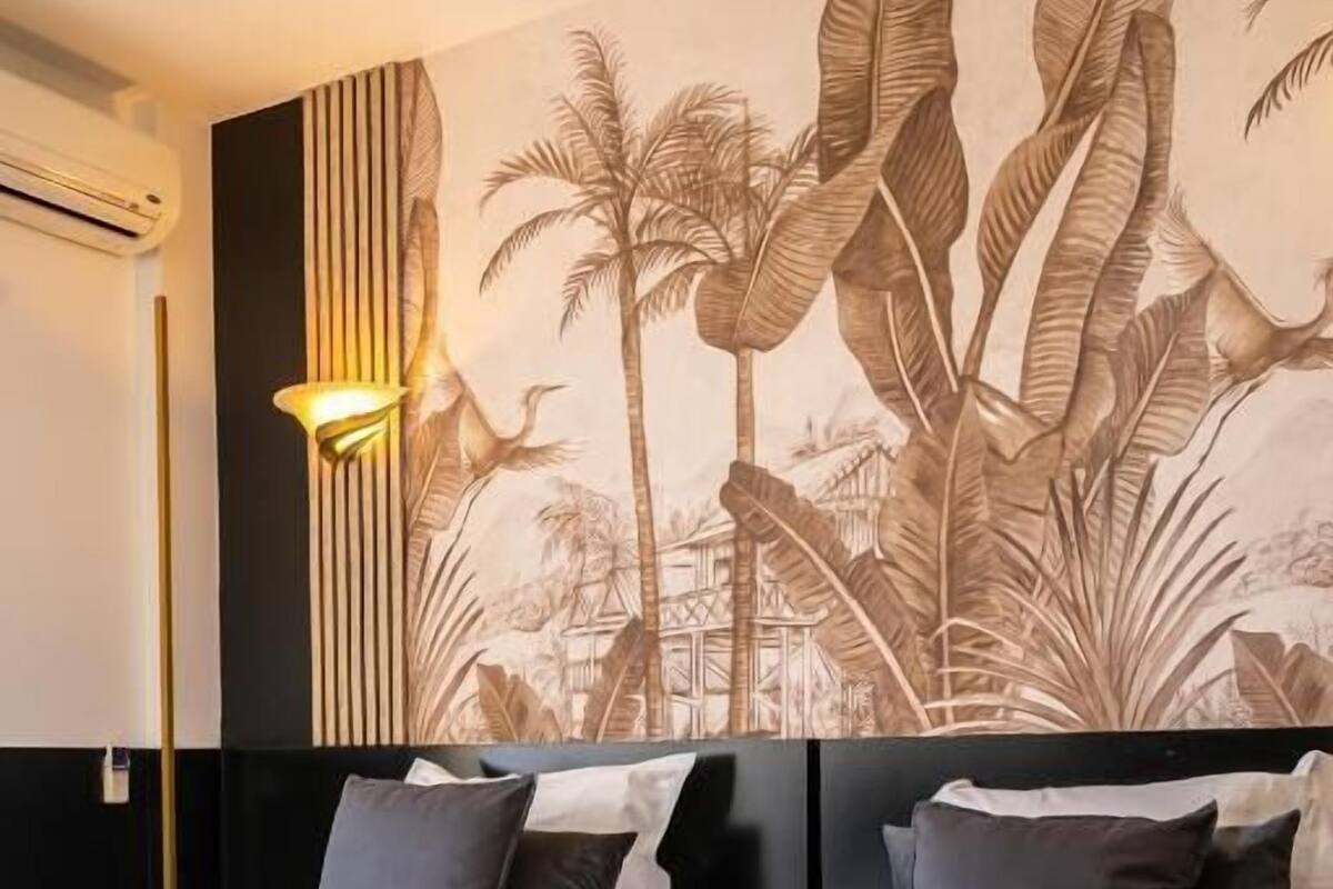 Quarto de hotel com duas camas, um grande mural na parede com plantas tropicais, uma mesinha de cabeceira com um porta-cosméticos e uma luminária moderna.