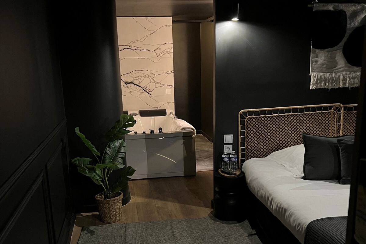 Um quarto com pouca luz, com uma cama, uma mesinha, um vaso de planta e uma parede com padrão semelhante ao mármore.