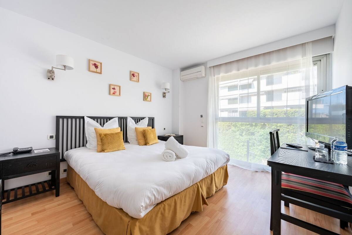 Quarto de hotel com cama, mesas de cabeceira, escrivaninha com cadeira, televisão e janela com vista.