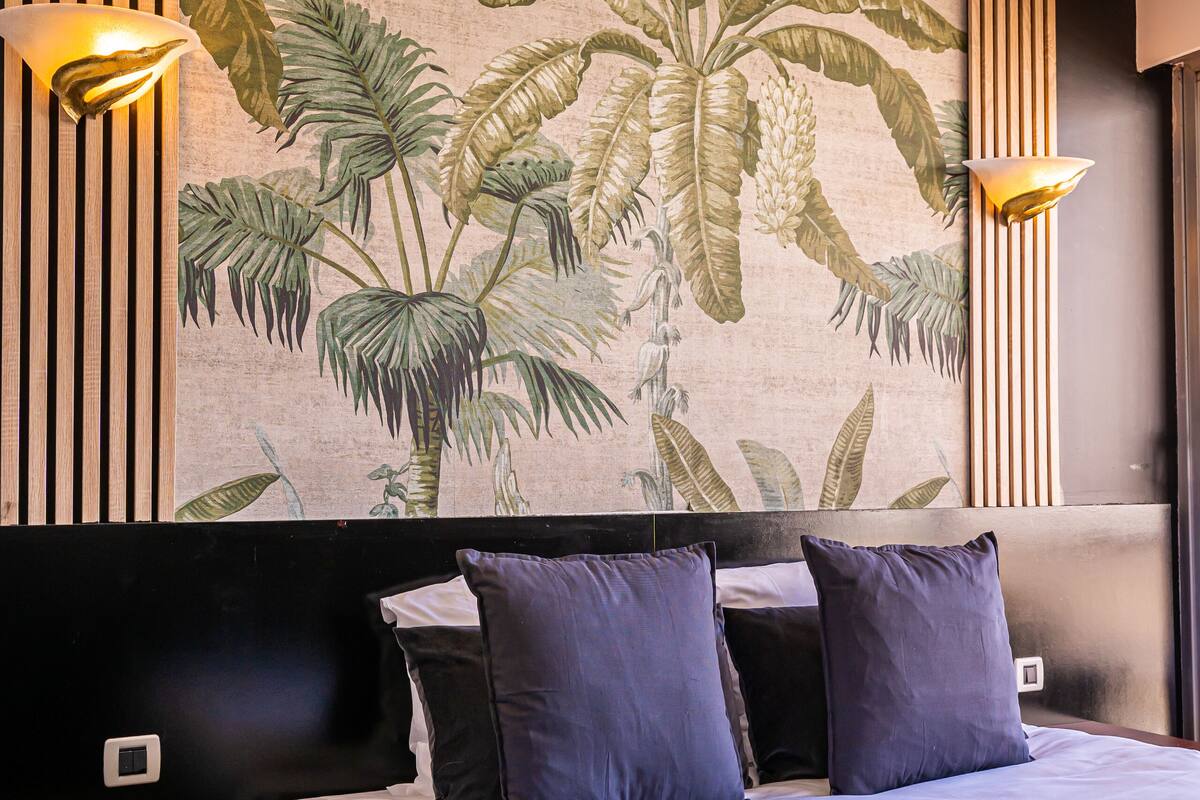 Quarto de hotel moderno com cama grande, cabeceira de madeira escura e parede com padrão de folhagem tropical.
