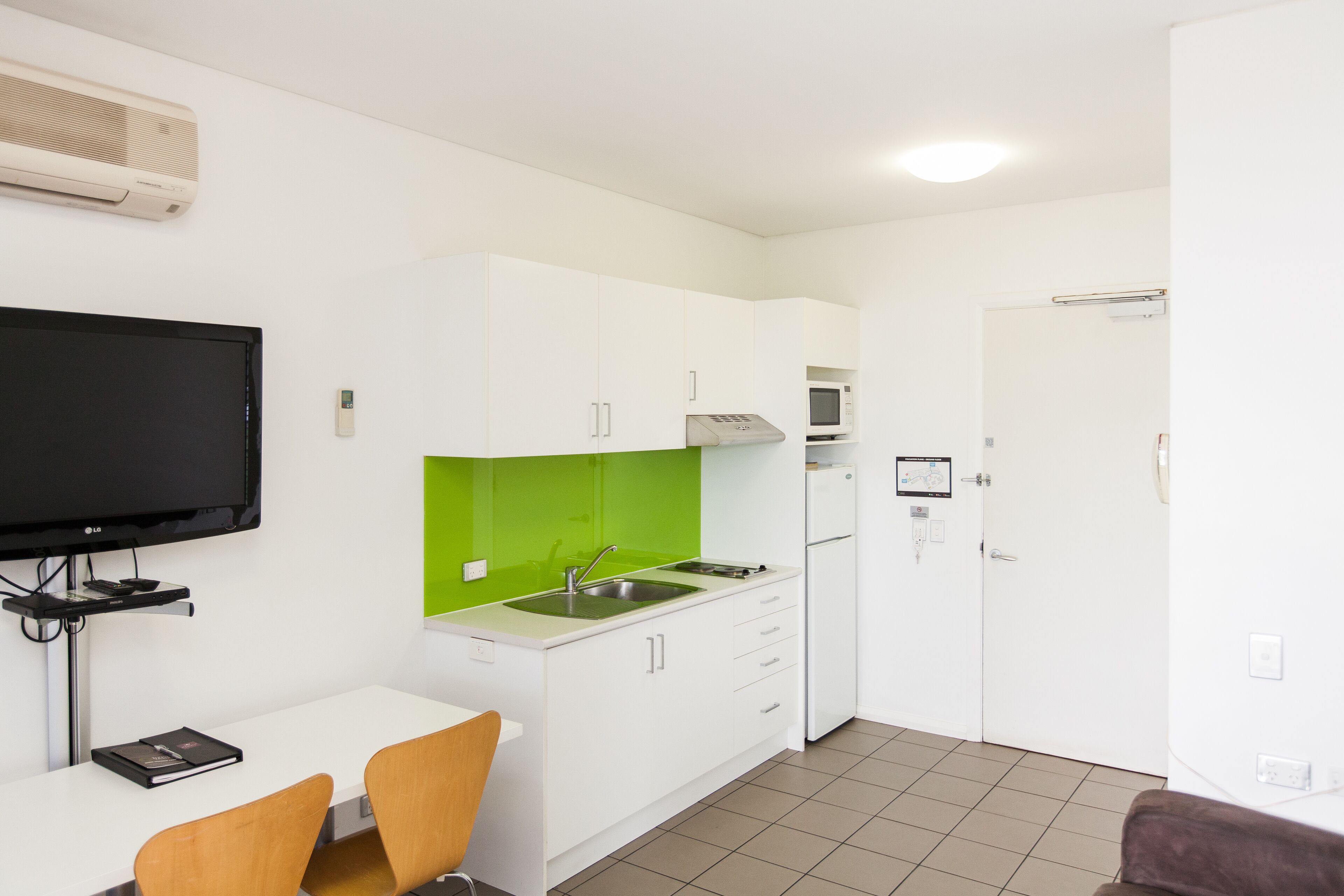 Cozinha compacta com armários brancos, painel de parede verde, pia, micro-ondas e geladeira. Há uma TV de tela plana presa na parede acima do balcão.