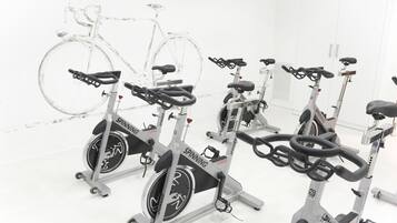 Várias bicicletas ergométricas alinhadas numa academia com paredes brancas e uma janela grande.