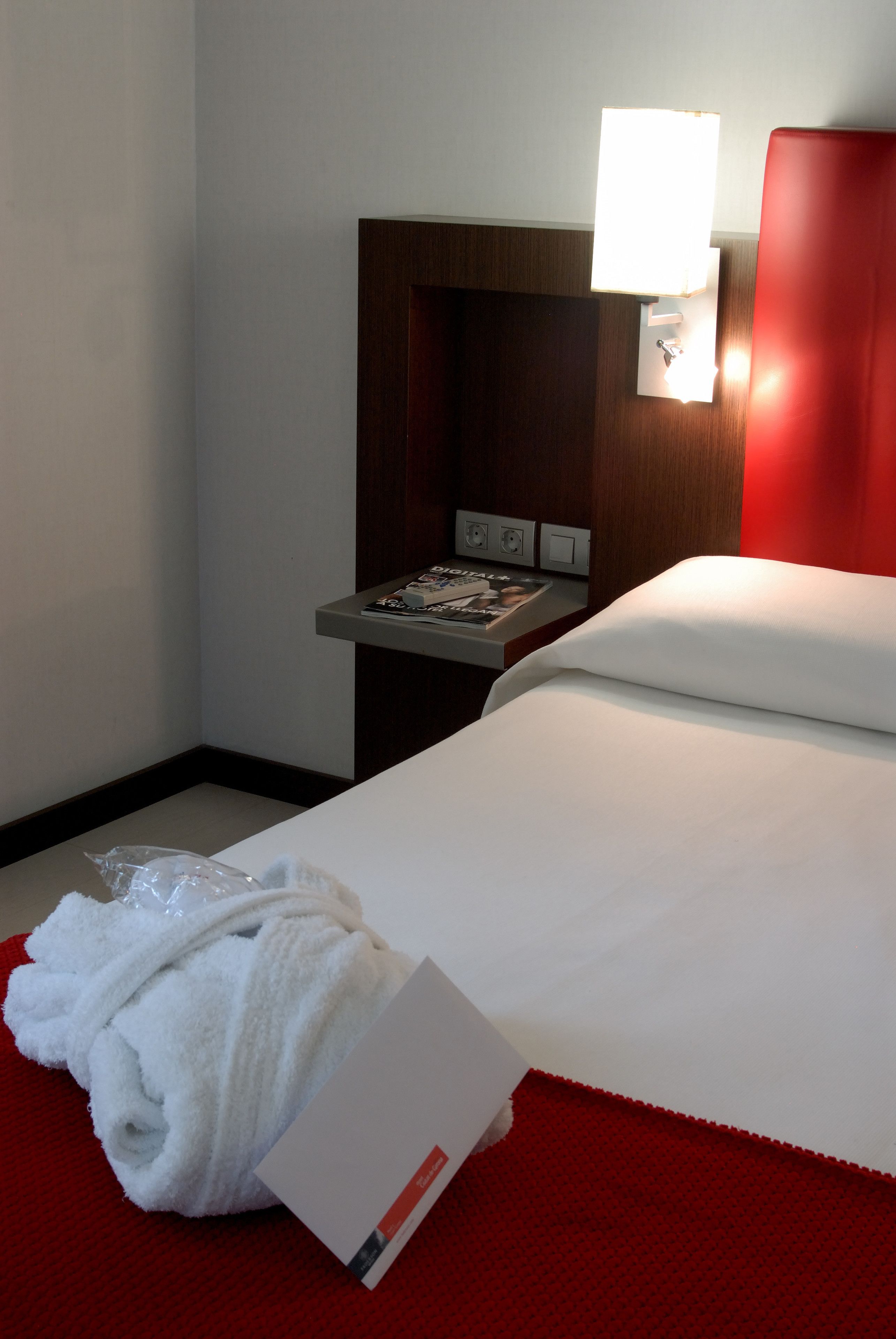 Quarto de hotel com uma cama bem arrumada, uma parede de destaque vermelha, uma mesa de cabeceira com abajur e uma toalha no chão.