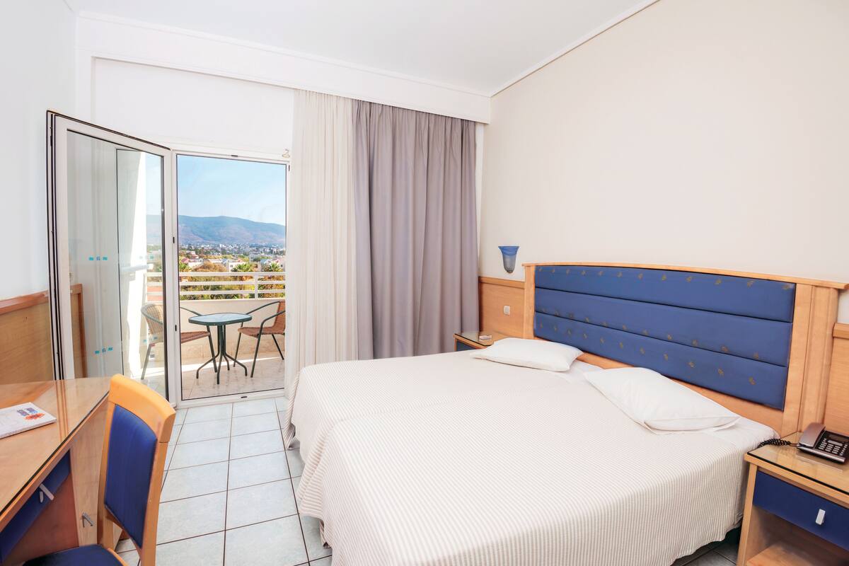 Quarto de hotel com cama, escrivaninha, cadeira, mesinha e vista da cidade por uma porta deslizante.