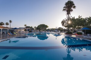 Uma piscina grande ao ar livre com várias pistas de natação, cercada por espreguiçadeiras e palmeiras.