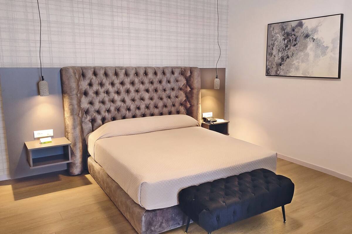 Quarto com cabeceira estofada, mesas de cabeceira, um banco e um quadro na parede.