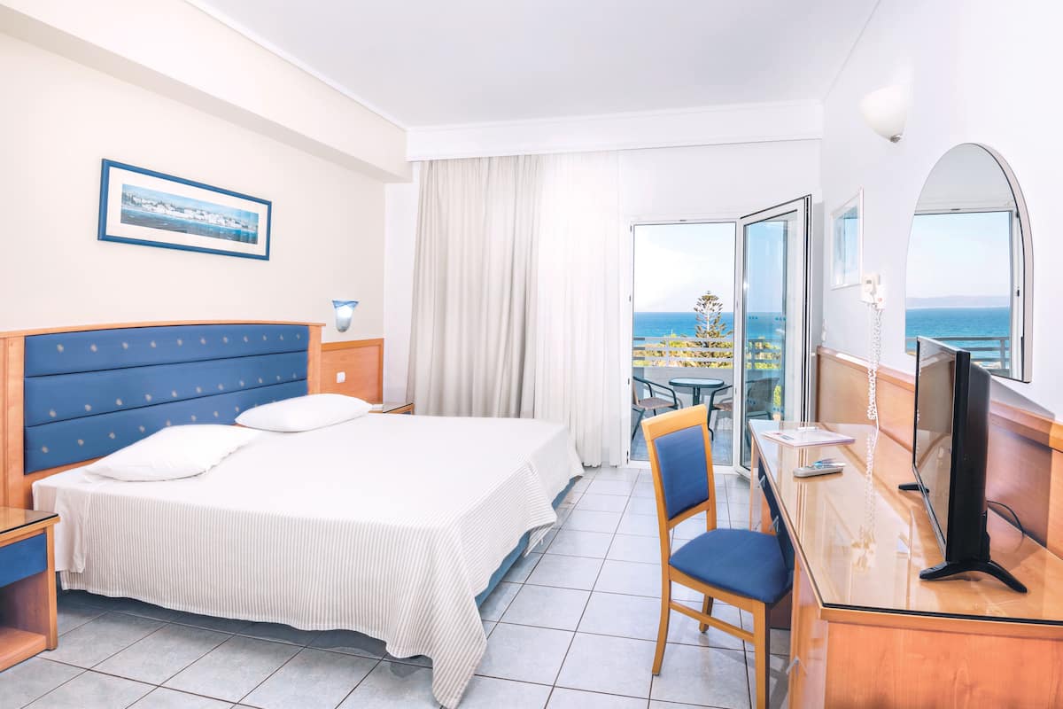 Quarto de hotel com cama, escrivaninha com computador, cadeira e vista para o mar.