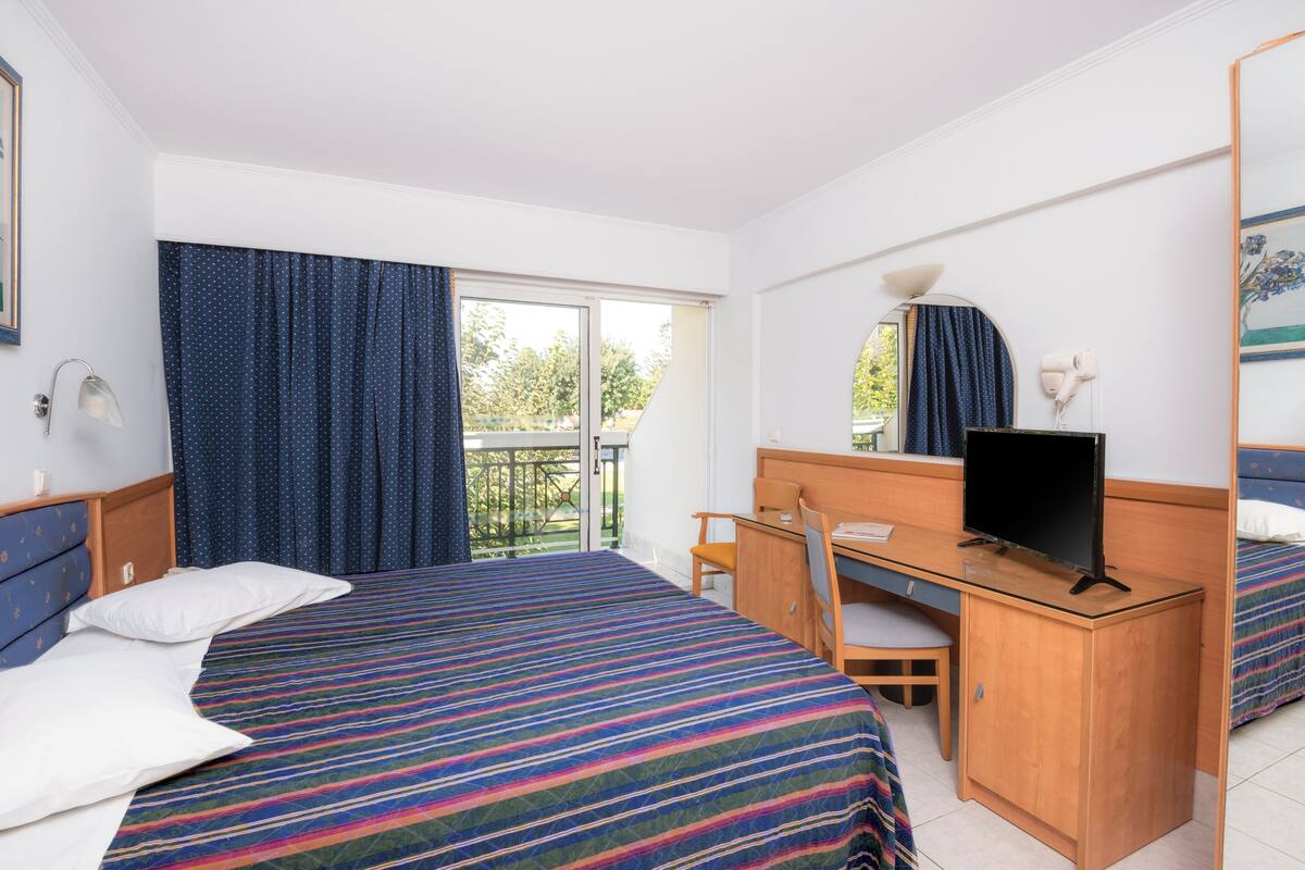 Quarto de hotel com cama, escrivaninha com computador, televisão, cadeira e janela com cortinas.