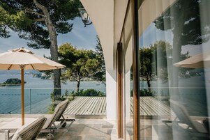 Uma varanda com deck de madeira, um guarda-sol bege e vista para o mar e árvores.